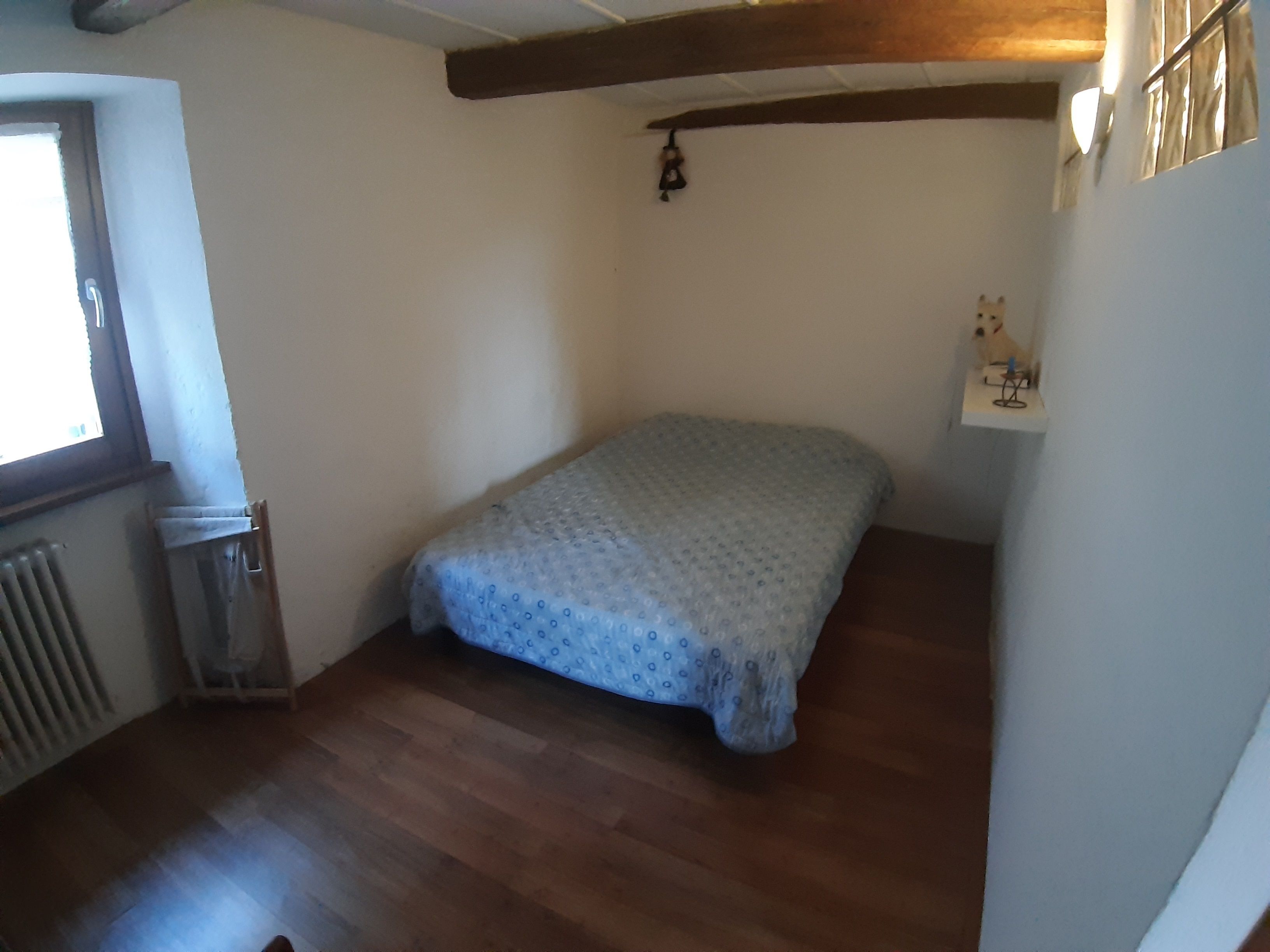 Apartamento pequeno e de um único quarto, com piso de madeira, uma cama com colcha estampada, um radiador, uma pequena prateleira com uma estatueta e uma janela com vista para o exterior.
