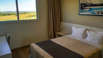 Quarto de hotel com cama, escrivaninha, janela com vista para o campo e um quadro na parede.