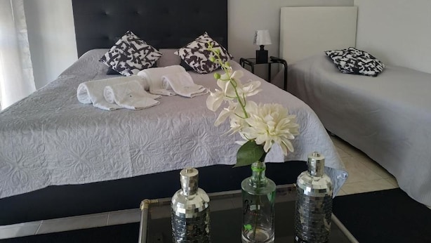 Cama bem arrumada com edredom branco, duas almofadas estampadas e um vaso com flores brancas. Há duas mesas de cabeceira, uma com abajur e a outra com uma garrafa.