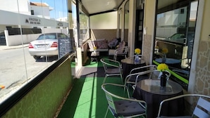 Uma área de estar coberta ao ar livre com piso verde, cadeiras de metal e mesas decoradas com flores amarelas.