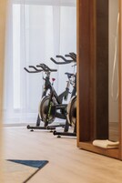 Quarto com duas bicicletas ergométricas, piso de madeira e uma janela grande com cortinas leves.