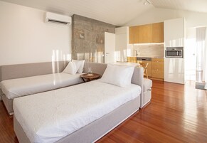 Quarto moderno com duas camas, uma pequena mesa de cabeceira e uma cozinha compacta ao fundo.