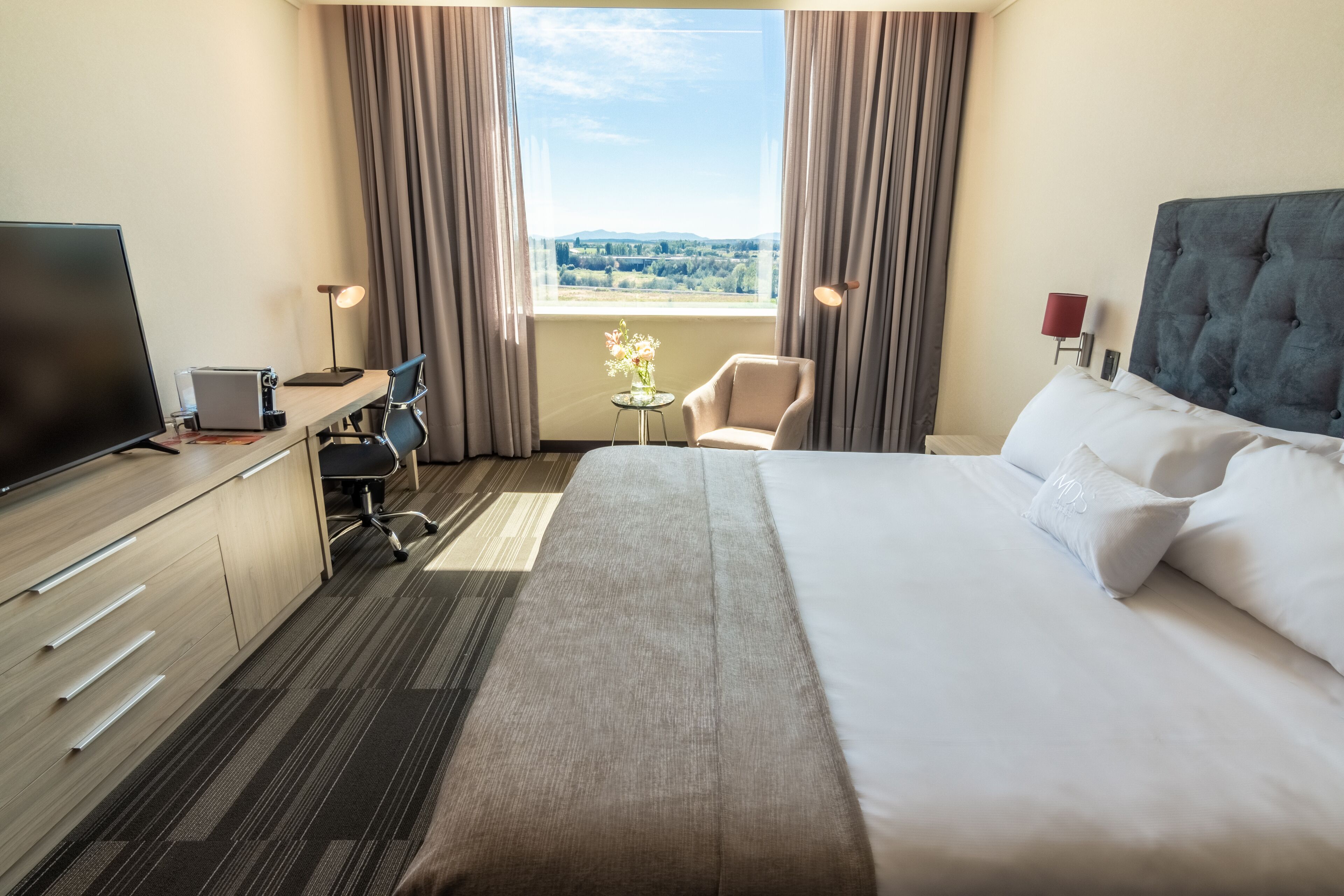 Quarto de hotel com uma cama grande, uma escrivaninha com computador, uma cadeira, uma TV e vista para o exterior.