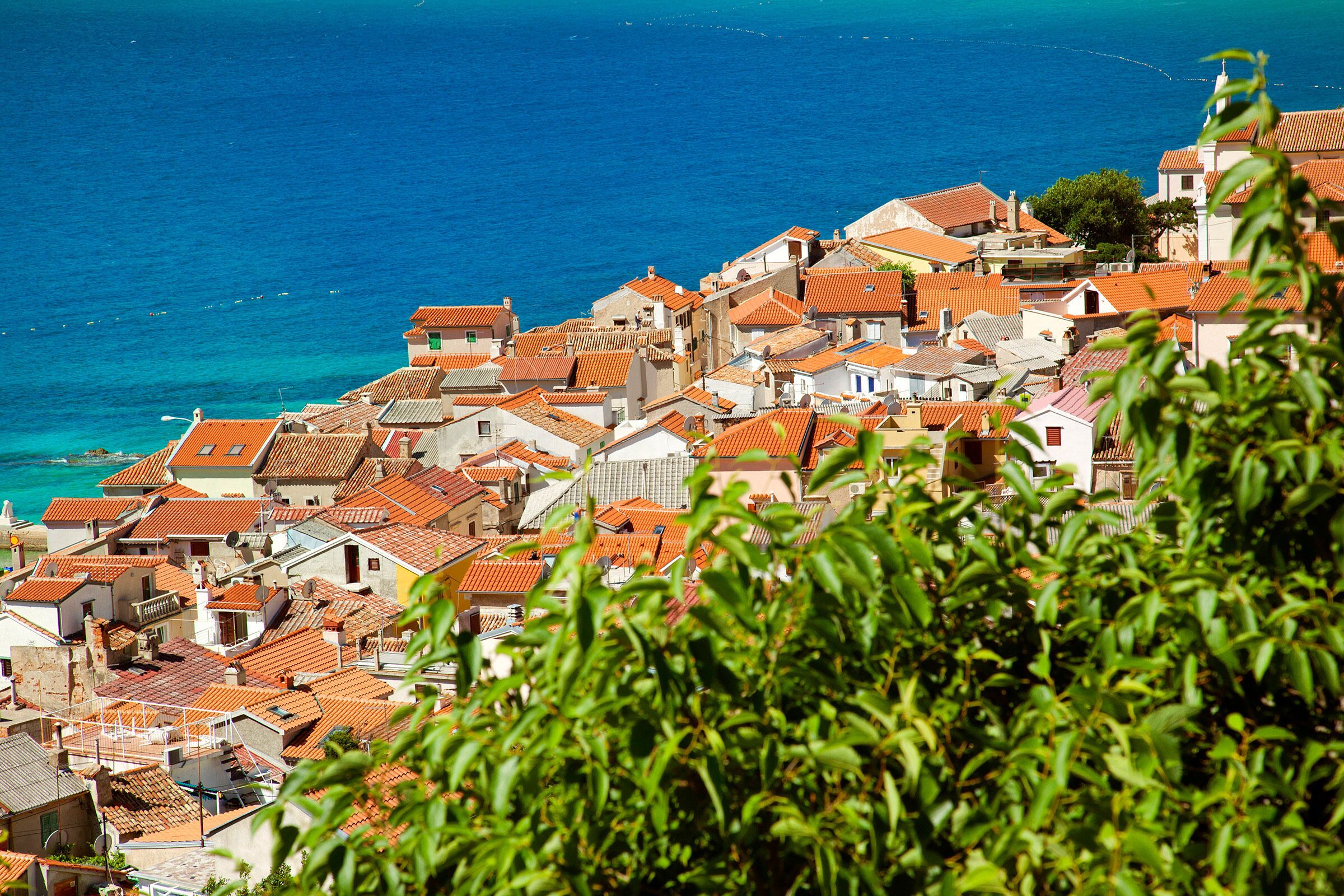 Uma vila costeira com edifícios de telhados em tons de terracota, mar azul cristalino e vegetação exuberante em primeiro plano.