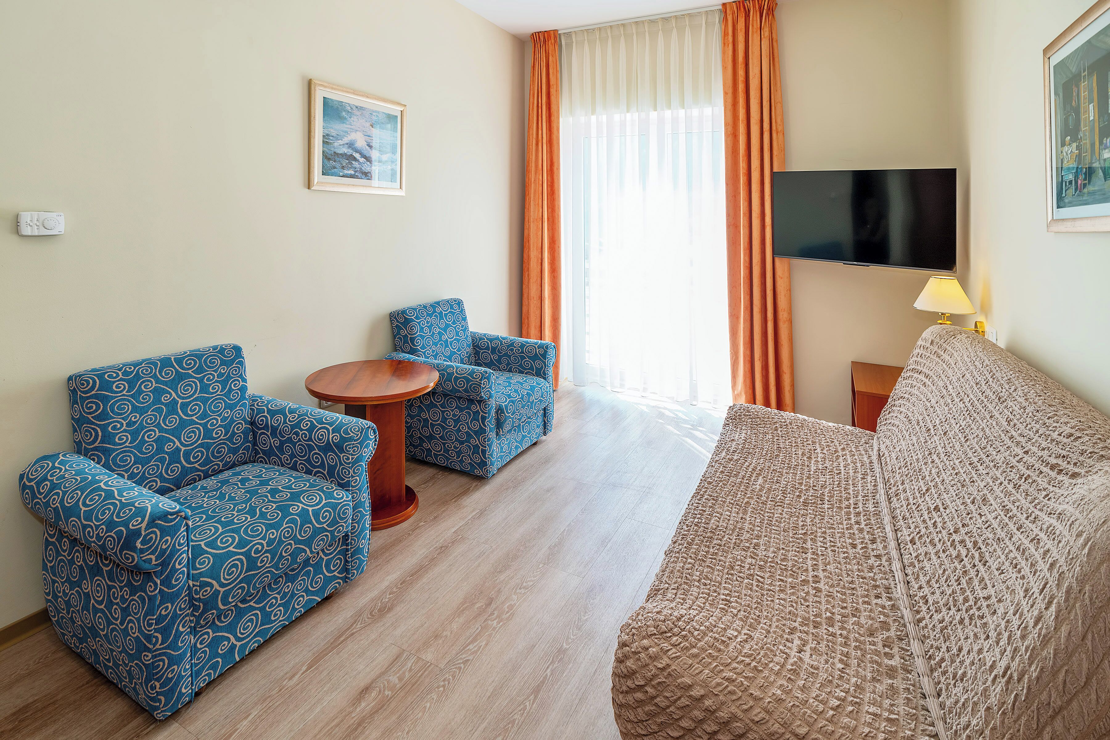 Quarto de hotel com sofá, duas poltronas estampadas, uma mesa redonda pequena, televisão e janela com cortinas.