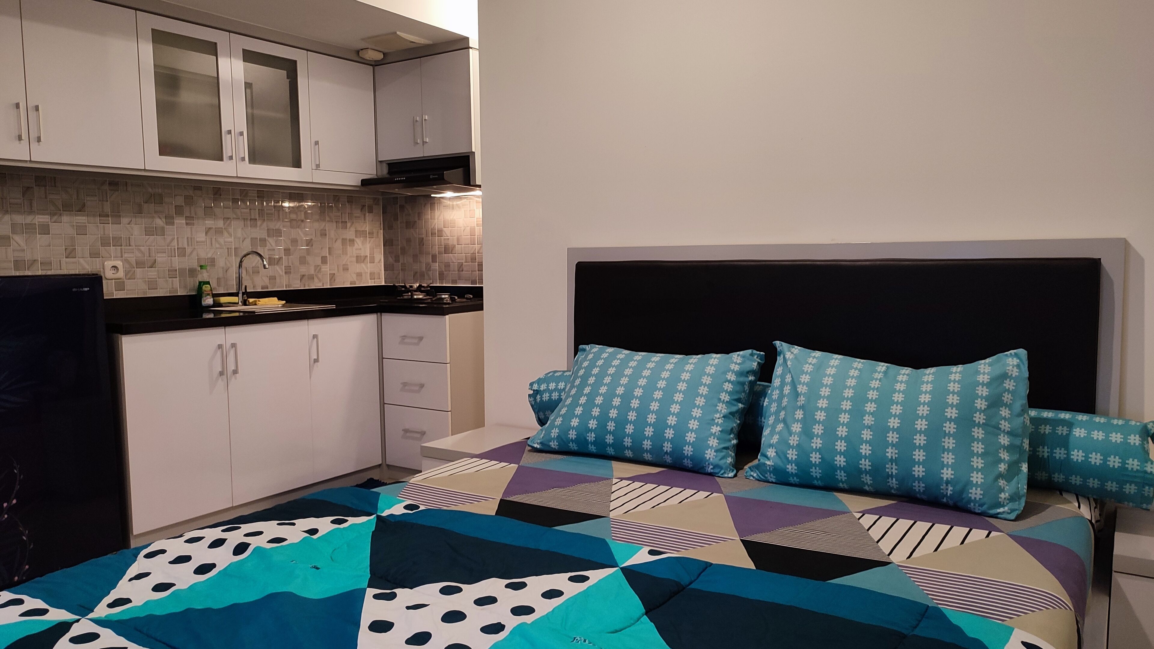 Quarto com cama, travesseiros e colcha. Há uma área de cozinha com armários e uma geladeira.
