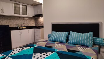 Quarto com cama, travesseiros e colcha. Há uma área de cozinha com armários e uma geladeira.