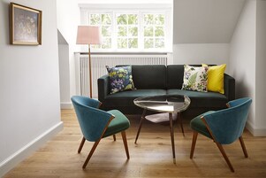 Uma sala de estar aconchegante com um sofá verde escuro, duas poltronas azul-esverdeadas, uma pequena mesa de centro redonda e um abajur.