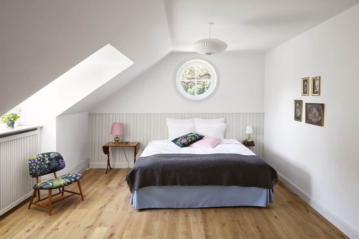 Quarto com teto inclinado, janela redonda, cama com roupa branca, cadeira estampada com padrão floral e mesa de cabeceira pequena com abajur.