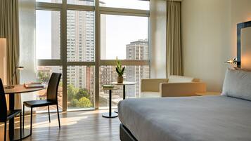 Quarto de hotel com uma cama grande, uma escrivaninha com cadeira, uma mesinha com uma planta e vista para edifícios altos pelas amplas janelas.
