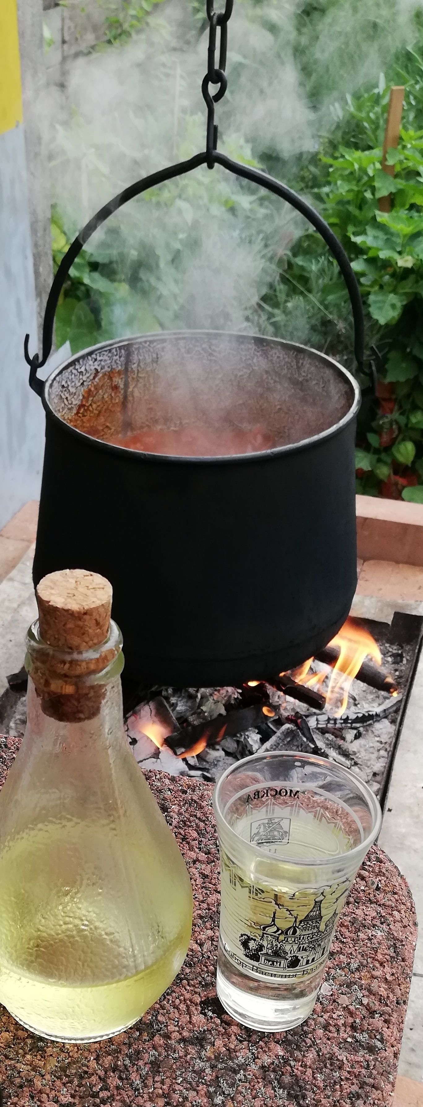 Uma panela preta em cima de um fogão, com vapor subindo, uma garrafa com rolha e um copo com etiqueta.