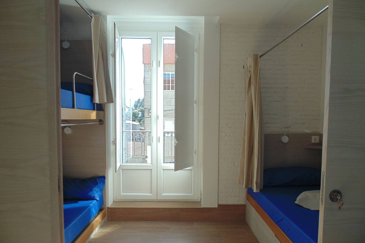 Quarto pequeno e compacto com piso de madeira, uma cama com colchão azul, uma cama beliche com colchão azul e uma porta branca que dá para uma varanda externa.