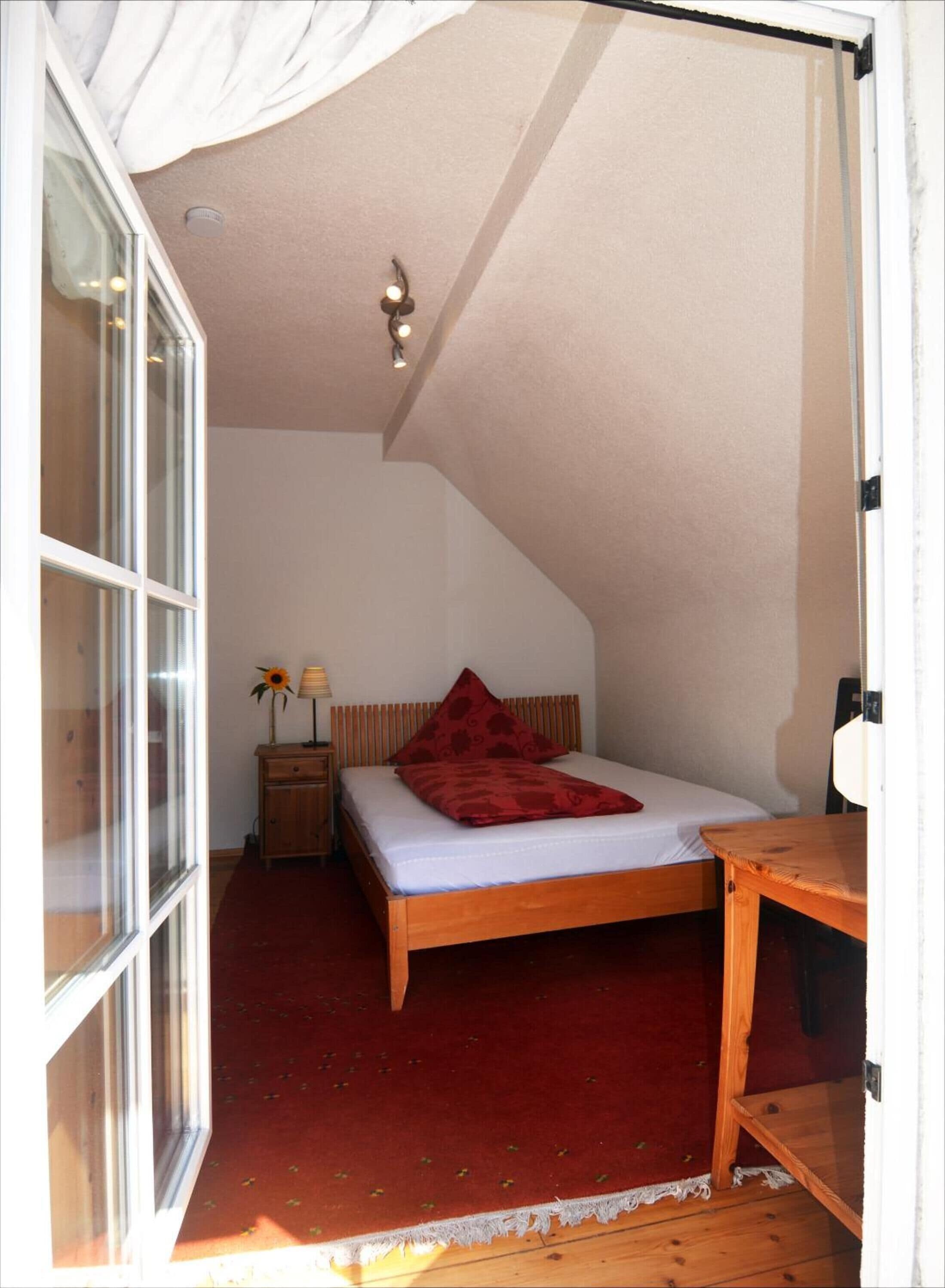 Um quarto pequeno e acolhedor com teto inclinado, uma cama de solteiro com manta vermelha, uma mesa de cabeceira com abajur e uma janela com cortinas.