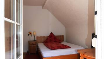 Um quarto pequeno e acolhedor com teto inclinado, uma cama de solteiro com manta vermelha, uma mesa de cabeceira com abajur e uma janela com cortinas.