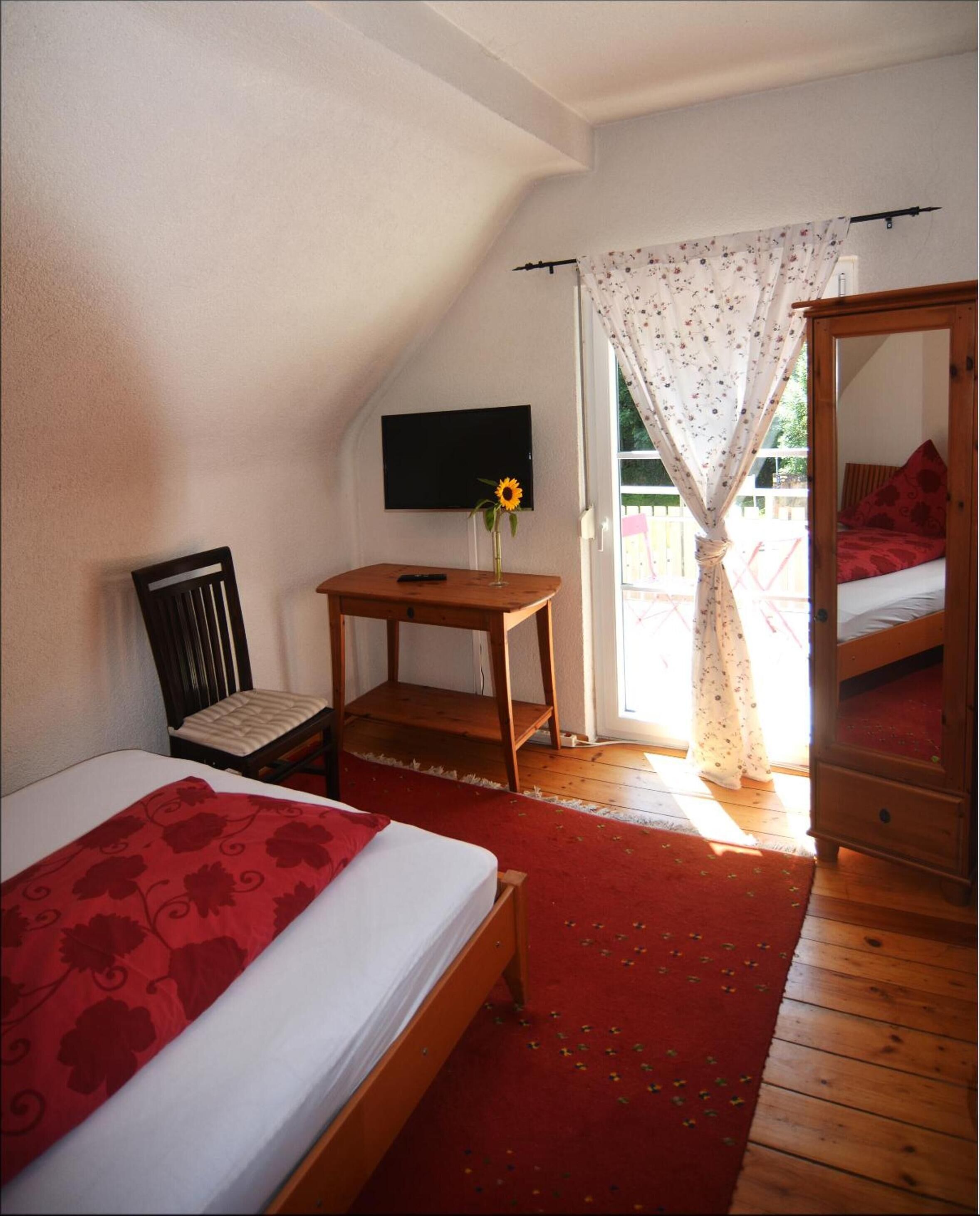 Quarto com uma cama, secretária de madeira, cadeira, televisão e janela com cortinas.