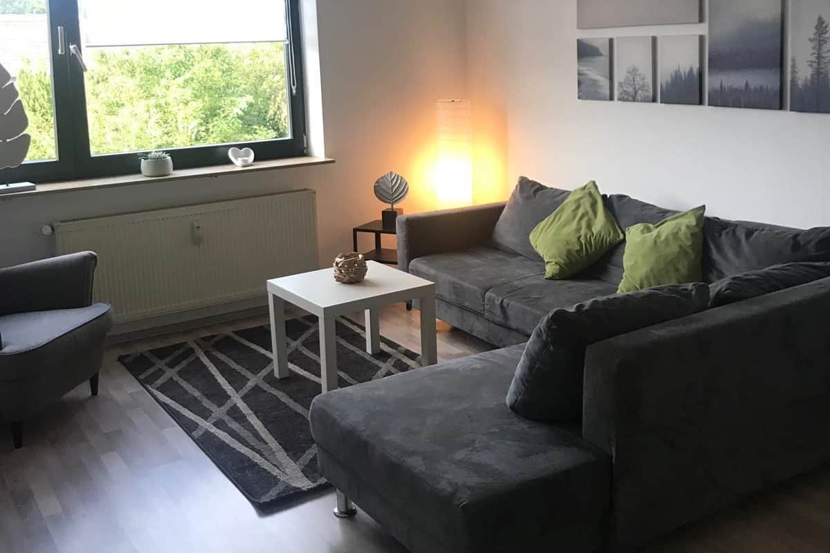 Uma sala de estar moderna com um sofá cinzento, uma mesa de centro branca e um abajur. Há uma janela grande que oferece luz natural e vista para o verde lá fora.