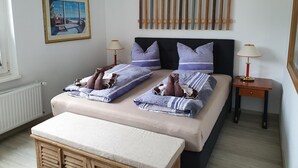 Quarto com uma cama, duas mesinhas de cabeceira, cabeceira com palhetas de madeira, um quadro emoldurado e uma janela com persianas.