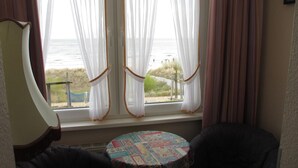 Quarto com vista para a praia através de cortinas translúcidas, uma pequena mesa redonda com toalha de mesa colorida e um sofá de cor escura.