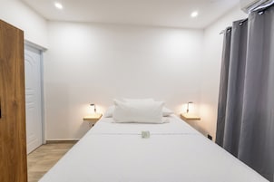 Uma cama de solteiro com colchão e almofadas brancas, com duas mesas de cabeceira com luminárias, em um quarto com porta de madeira e cortina cinza.