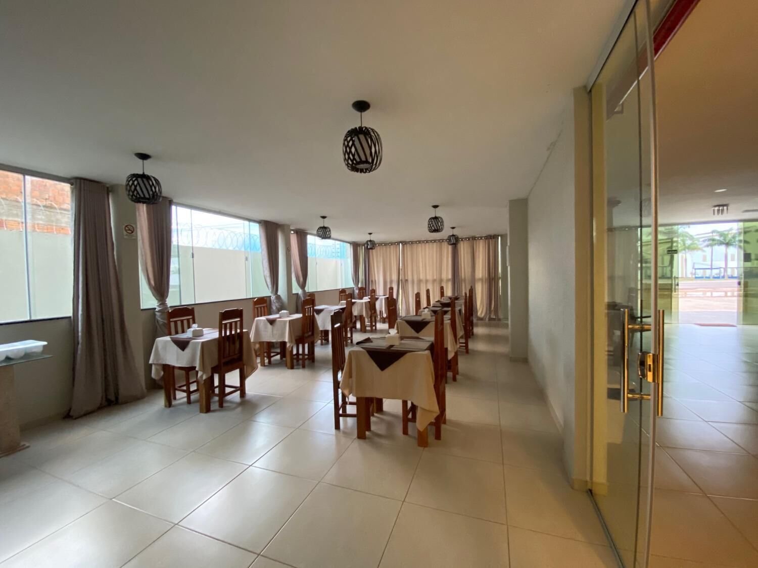 Uma sala de jantar com mesas e cadeiras, grandes janelas com cortinas e luminárias pendentes.