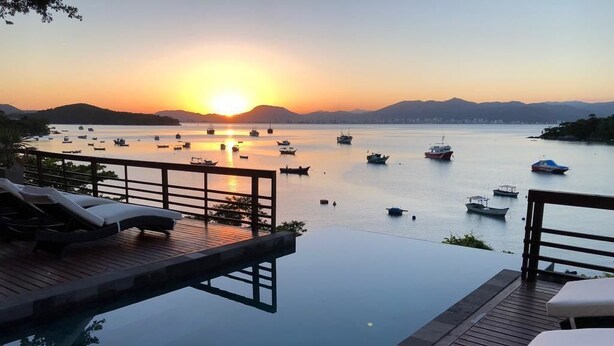 Piscina infinita com espreguiçadeiras, um deck de madeira e vista para barcos em águas calmas ao pôr do sol.