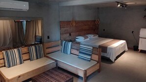 Quarto com sofá, duas camas, cabeceira de madeira, uma mesinha e ar-condicionado fixado na parede.