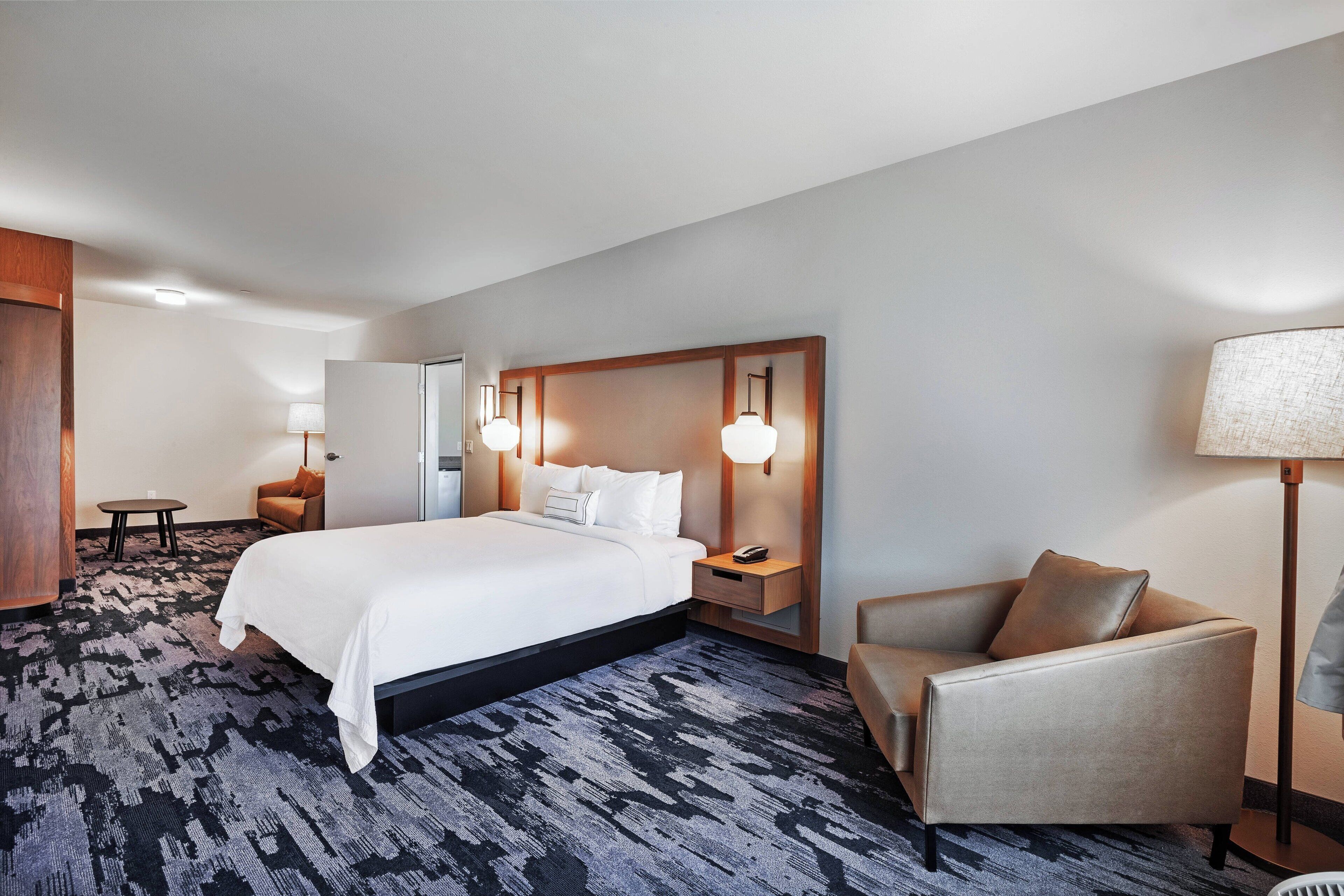 Quarto de hotel com uma cama grande, um sofá, uma mesa de cabeceira com abajur e piso carpeteado.