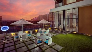 Uma área externa com mesas e cadeiras, uma placa com a inscrição "Sky Lounge" e vista para a cidade ao pôr do sol.