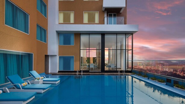 Piscina no terraço com espreguiçadeiras azuis, área de refeições envidraçada e vista para a cidade ao pôr do sol.