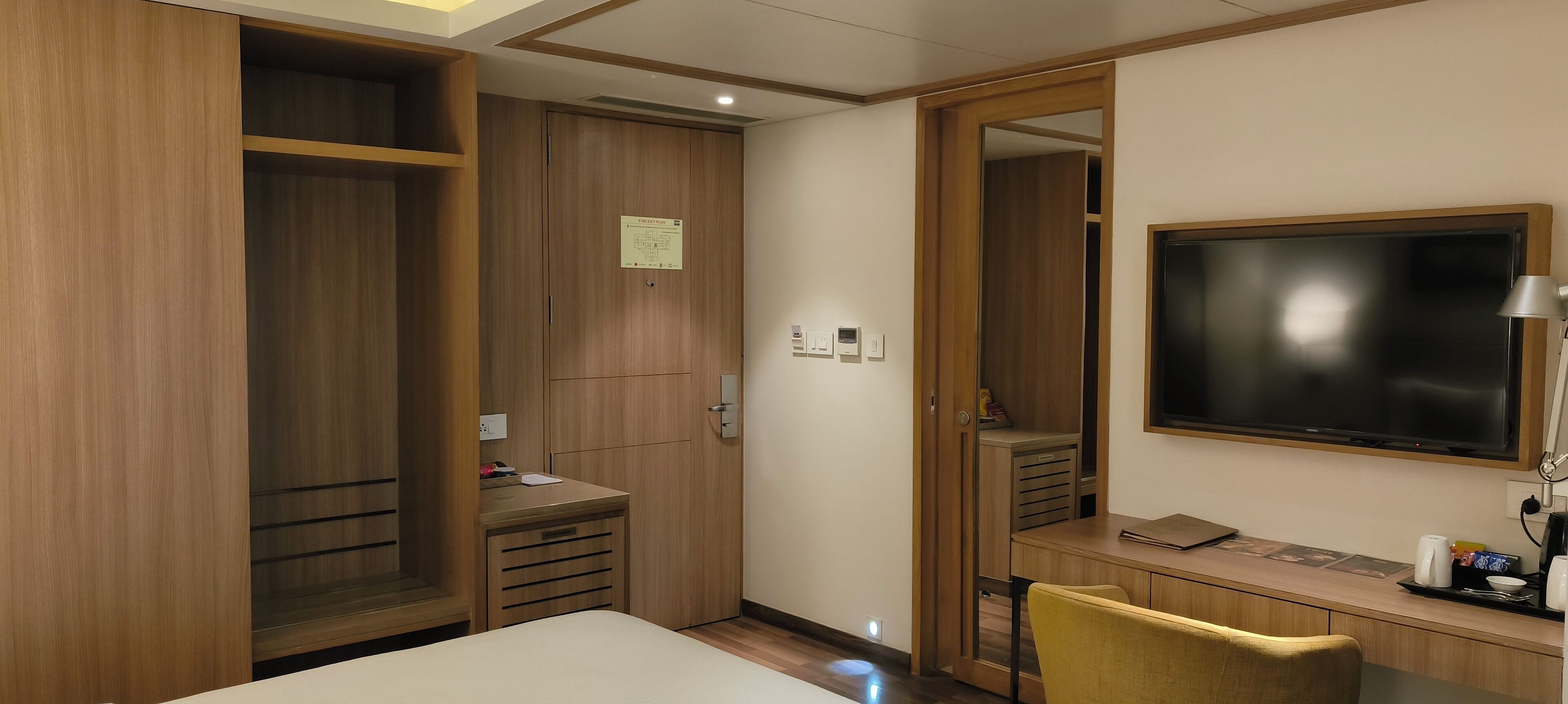 Quarto de hotel com guarda-roupa de madeira, escrivaninha com cadeira, televisão de tela plana e espelho.