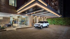 Entrada moderna de hotel com um SUV branco estacionado em frente, fachada de vidro e um sebe verde.