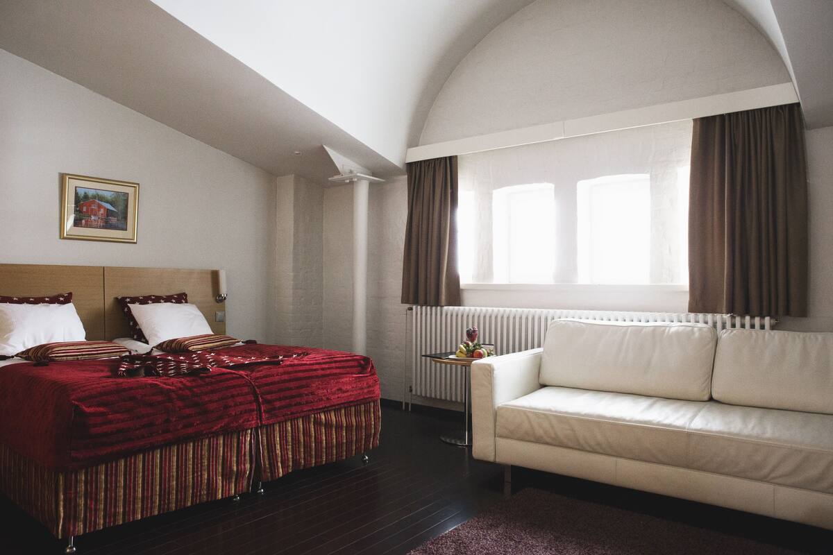 Quarto de hotel com cama, sofá, radiador, um quadro na parede e janela com cortinas.