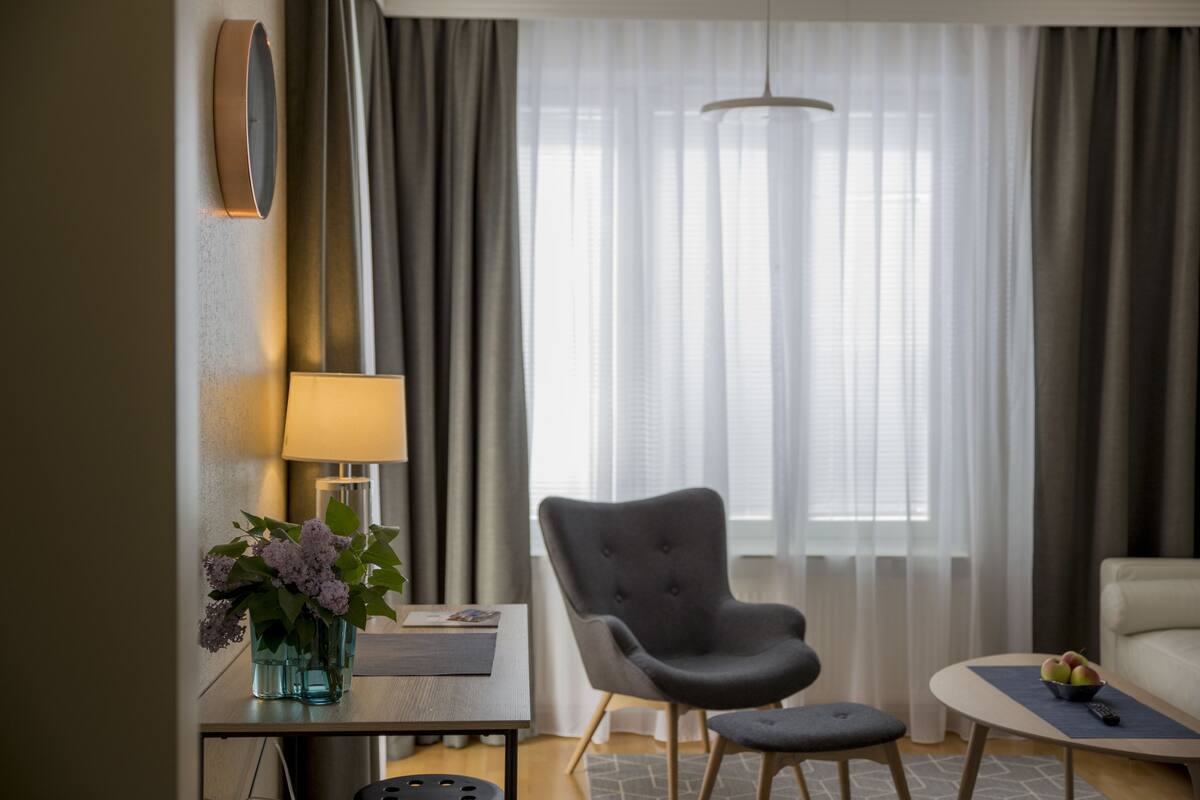 Quarto de hotel com uma cadeira moderna, uma pequena mesa com abajur, um vaso de flores e vista do exterior através de cortinas translúcidas.
