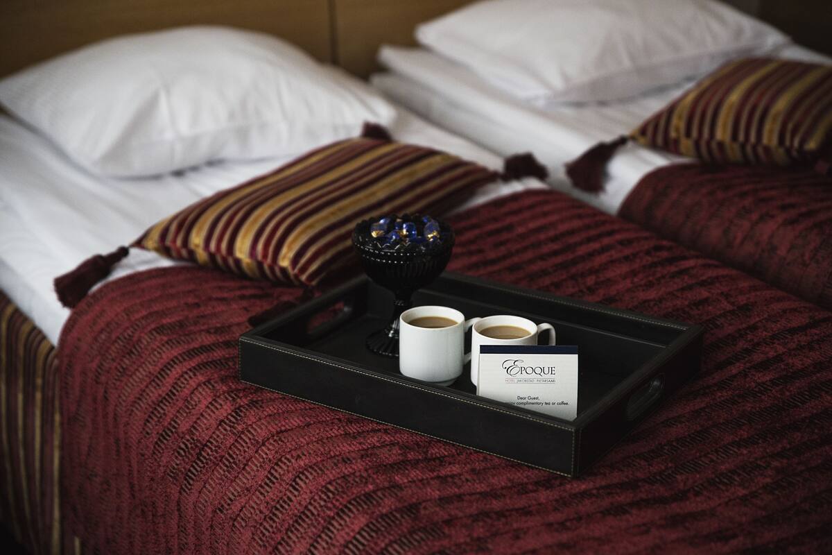 Quarto de hotel com duas camas, cada uma com almofadas brancas e cobertores vermelhos. Há uma bandeja com duas chávenas de café e uma pequena tigela decorativa.