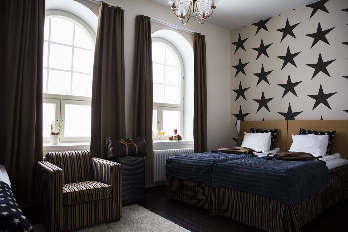 Quarto de hotel com uma cama grande, uma cadeira, janela com cortinas e parede com padrão de estrelas.