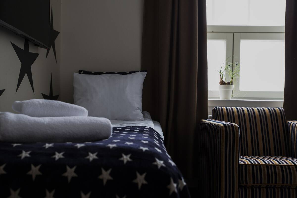 Quarto de hotel com cama, poltrona listrada, planta em vaso perto da janela e adesivos de estrelas na parede.