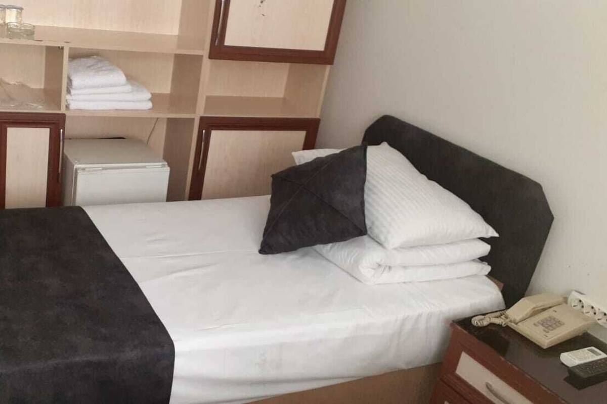 Quarto de hotel com uma cama, uma mesinha de cabeceira com telefone, uma prateleira com toalhas dobradas e um pequeno frigorífico.