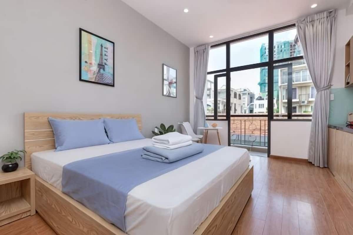 Quarto de hotel moderno com uma cama grande, uma pequena mesa de cabeceira, um criado-mudo, uma janela com vista para edifícios e uma varanda.