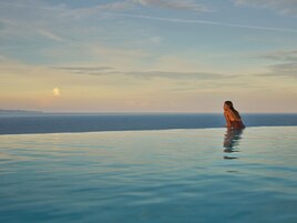 Uma mulher está em uma piscina infinita com vista para o oceano e um pôr do sol.
