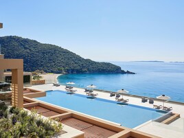 Uma área à beira da piscina com espreguiçadeiras e guarda-sóis, com vista para um mar azul cristalino e uma encosta arborizada.