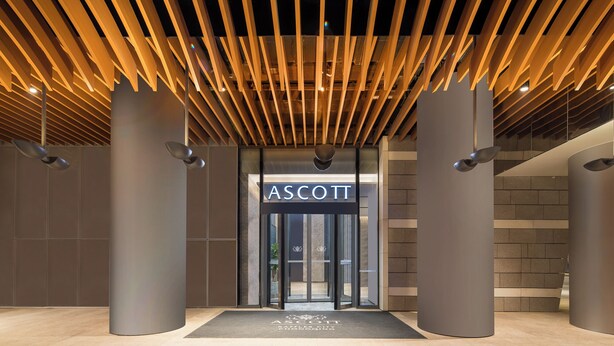 Uma entrada moderna com teto de ripas de madeira, porta de vidro e uma placa com o nome 'ASCOTT'.