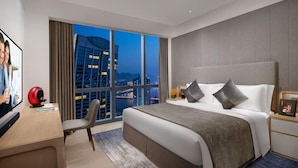 Quarto de hotel moderno com uma cama grande, uma escrivaninha e vista para a paisagem urbana ao entardecer.