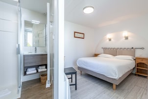 Quarto moderno com cama, mesas de cabeceira, um criado-mudo, uma pequena mesa, uma cadeira, prateleira com toalhas, espelho e luminária fixada na parede.