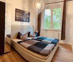 Um quarto com cama, pintura de paisagem urbana, lustre, janela com cortinas e radiador.