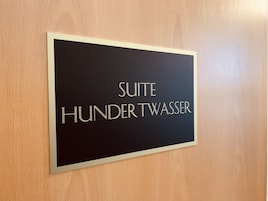 Uma placa com o texto "SUITE HUNDERTWASSER" em fundo preto com borda branca, fixada em uma parede de madeira.