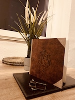 Um livro encadernado em couro marrom com texto em relevo, sobre uma capa de couro preta, ao lado de um vaso com planta de folhas verdes longas e flores brancas, em uma superfície de madeira perto de uma janela.