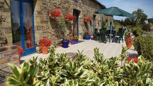 Um edifício de pedra com porta azul e janelas, área de estar externa com cadeiras e mesas, e vasos com plantas e flores vermelhas.