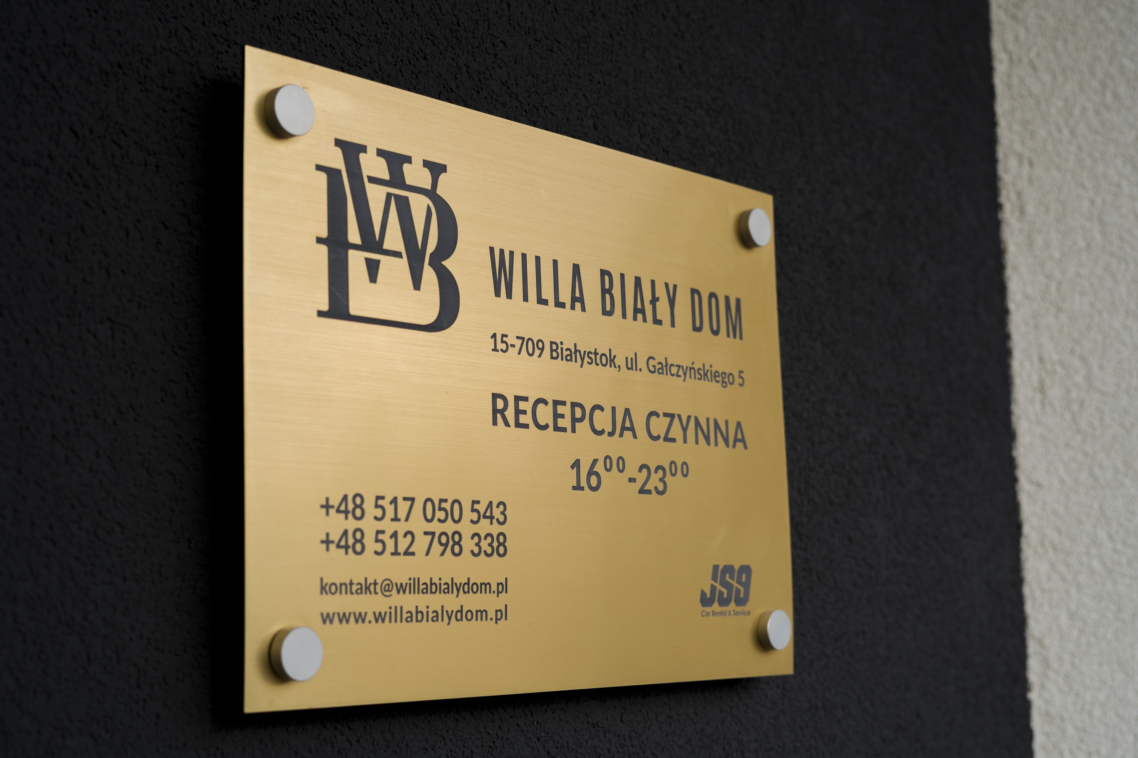 Uma placa com o nome "WILLA BIATY DOM" e informações de contacto, incluindo horário de funcionamento da receção das 16:00-23:00.