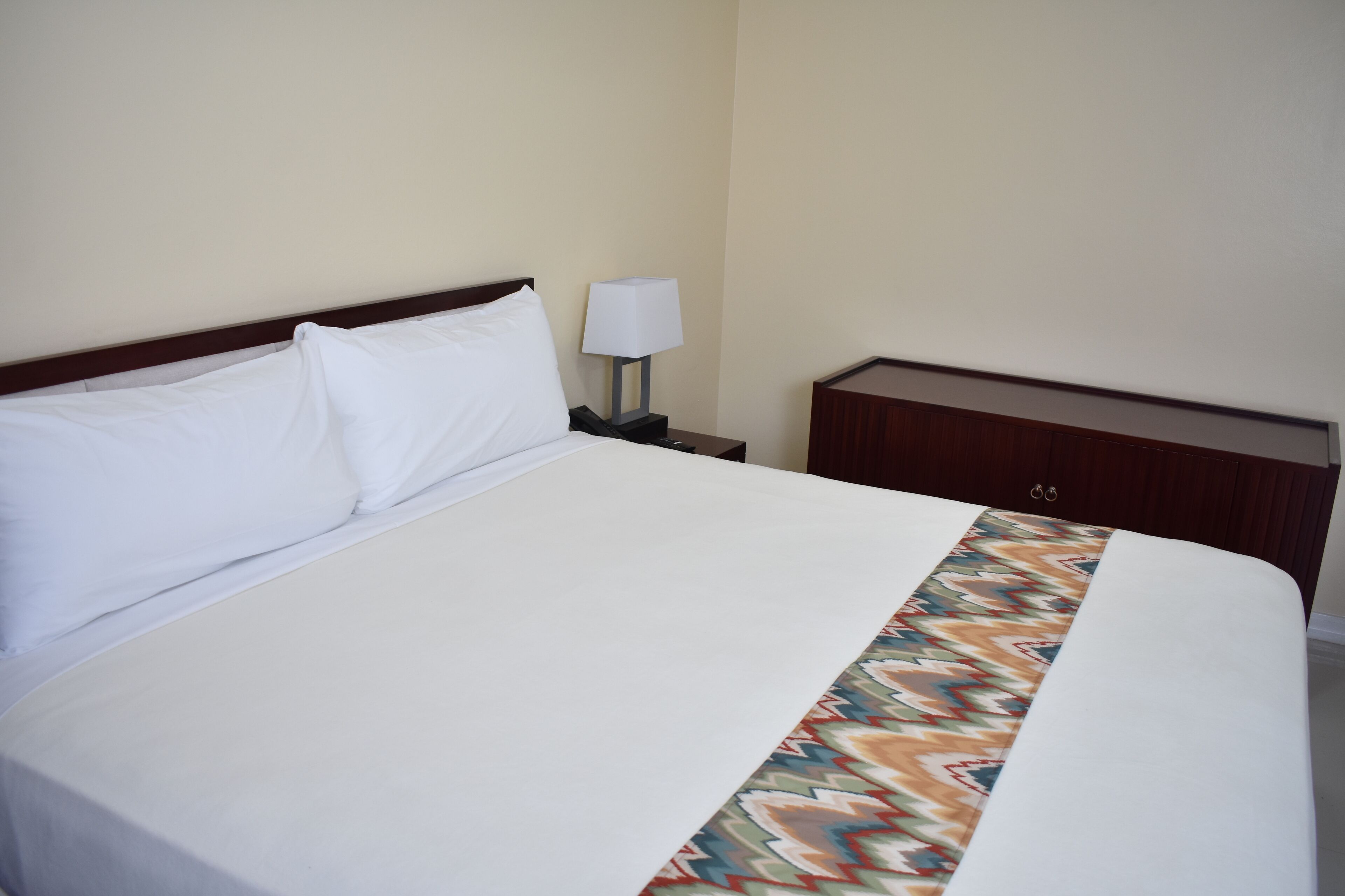 Cama bem arrumada com lençóis brancos e um corredor estampado, uma luminária de cabeceira e uma mesa de cabeceira de madeira.