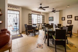 Uma sala de jantar com uma mesa grande e cadeiras, ventiladores de teto e vista pela janela com grades.